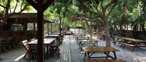 Restaurante ao ar livre com mesas e bancos de madeira, rodeado por árvores e coberto por uma pérgola de madeira.