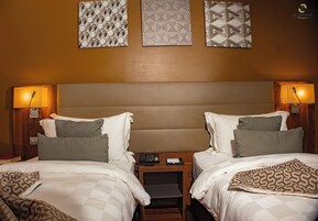 Quarto de hotel com duas camas, mesa de cabeceira com telefone e quadros nas paredes acima das camas.