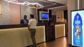 Uma área de recepção de hotel com design moderno, um balcão de atendimento, uma tela de televisão e um painel digital anunciando um restaurante.