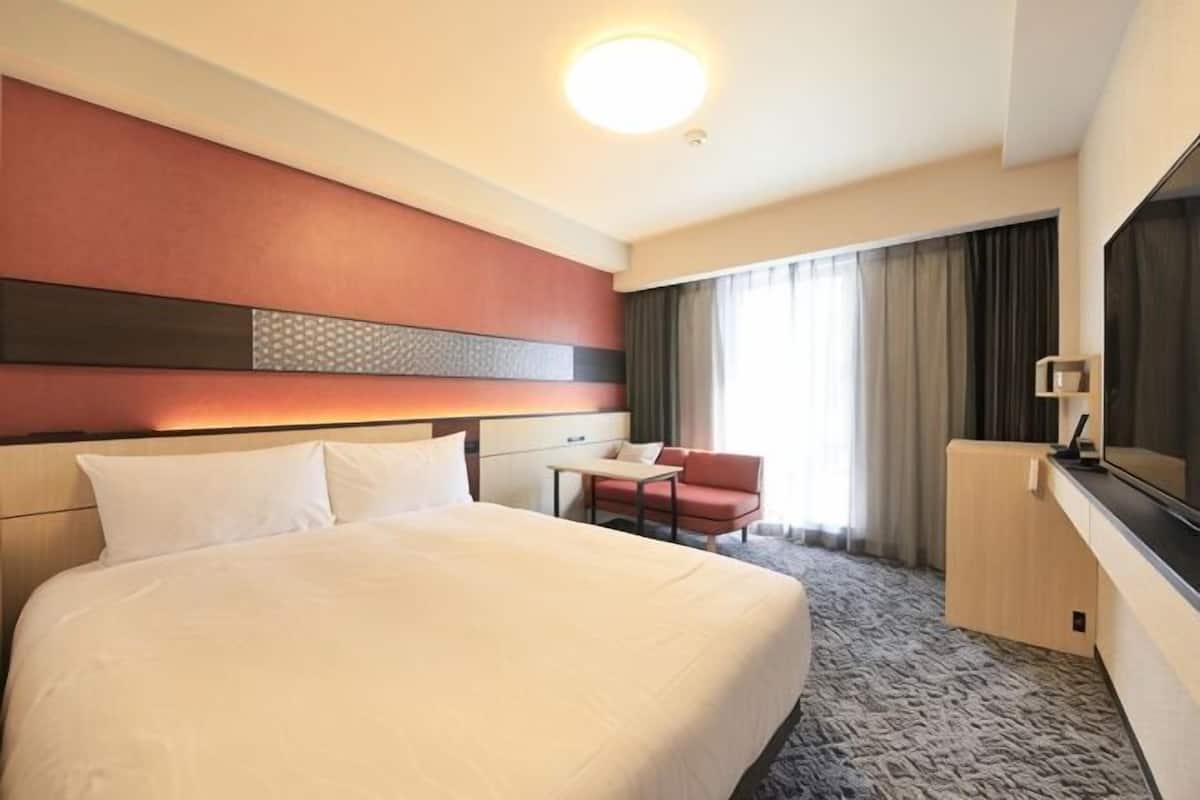 Quarto de hotel com uma cama grande, uma escrivaninha, uma cadeira, uma televisão e uma janela com cortinas.