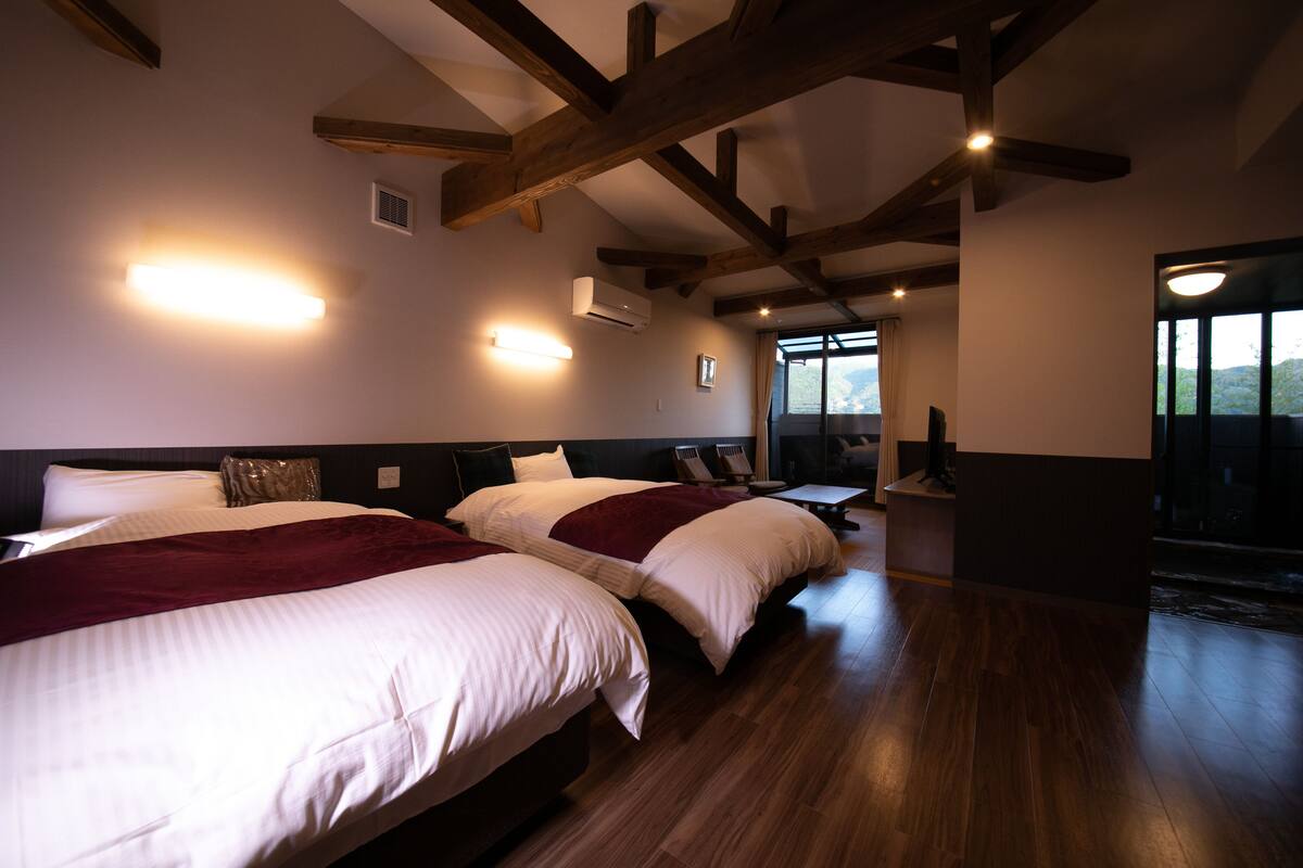 Quarto de hotel com duas camas grandes, vigas de madeira no teto, uma mesinha e vista pela janela.
