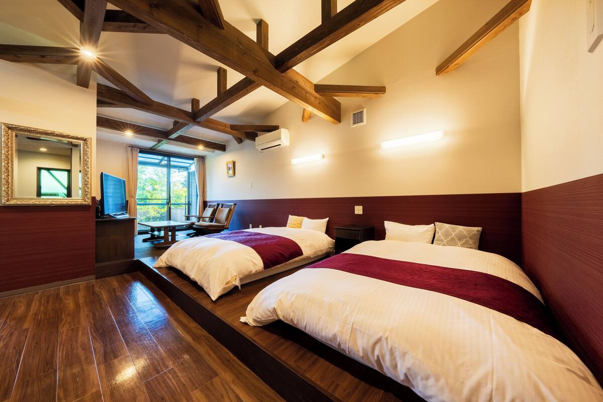 Quarto de hotel com duas camas, vigas de madeira no teto, um espelho grande e janela com vista para o verde.