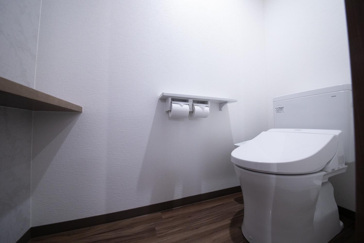 Casa de banho pequena e moderna com sanita, suporte de papel higiénico e prateleira de madeira.