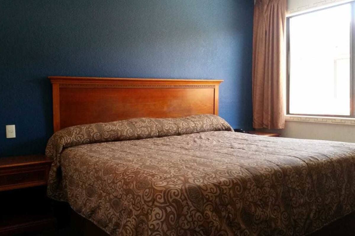 Quarto com cabeceira de madeira, colcha estampada, mesa de cabeceira e janela com cortinas.