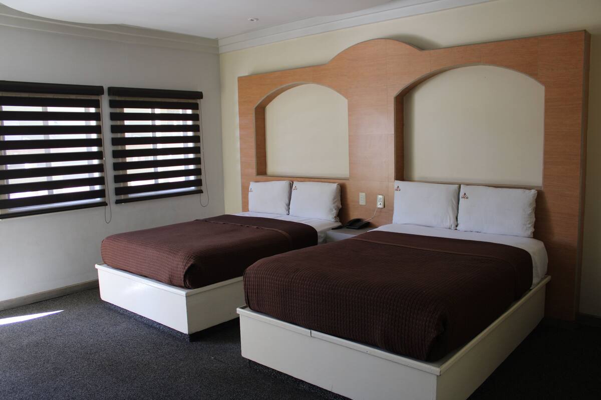 Quarto de hotel com duas camas de solteiro, cabeceira de madeira, mesa de cabeceira pequena e janela com persianas.