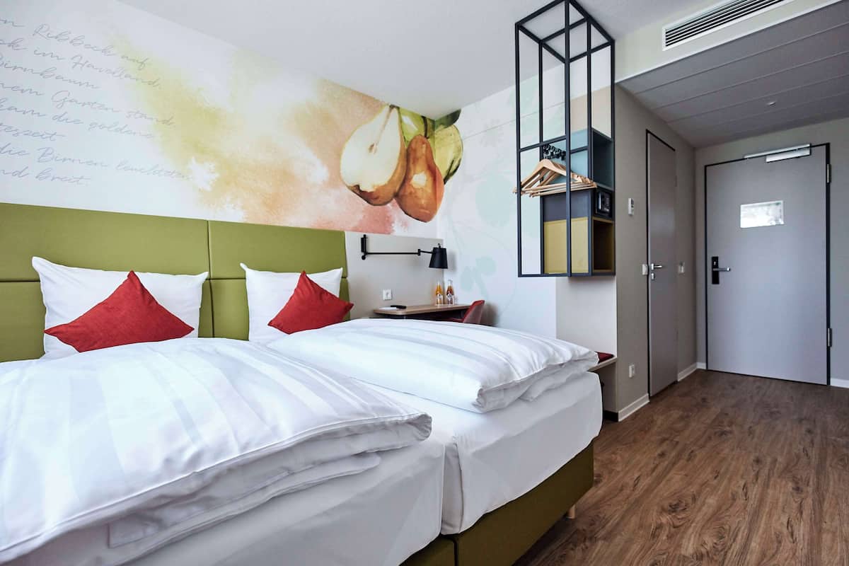 Quarto de hotel com cama, escrivaninha, cadeira, guarda-roupa e parede com pintura de frutas.