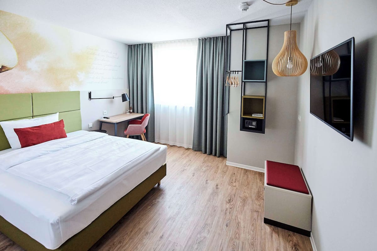 Quarto de hotel com cama, escrivaninha, cadeira e televisão na parede.