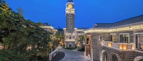 Um edifício tradicional com uma torre do relógio, rodeado por vegetação exuberante e um caminho pavimentado.