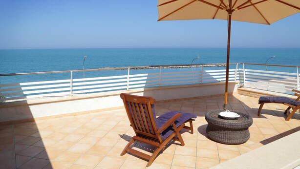 Um terraço com uma cadeira de madeira, um assento acolchoado, um banquinho de vime e um grande guarda-sol.