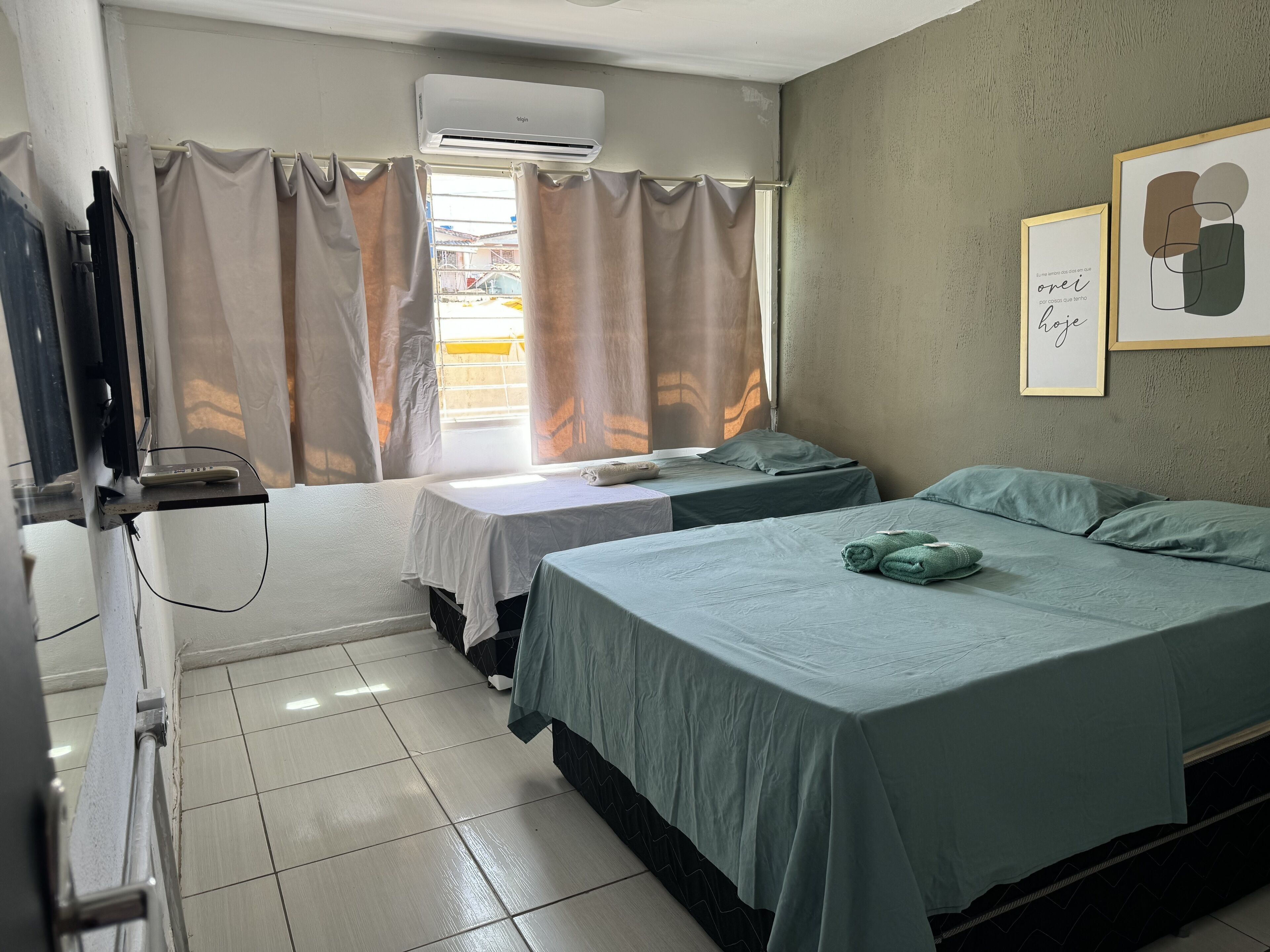 Quarto de hotel com duas camas, janela com cortinas, ar condicionado e televisão na parede.