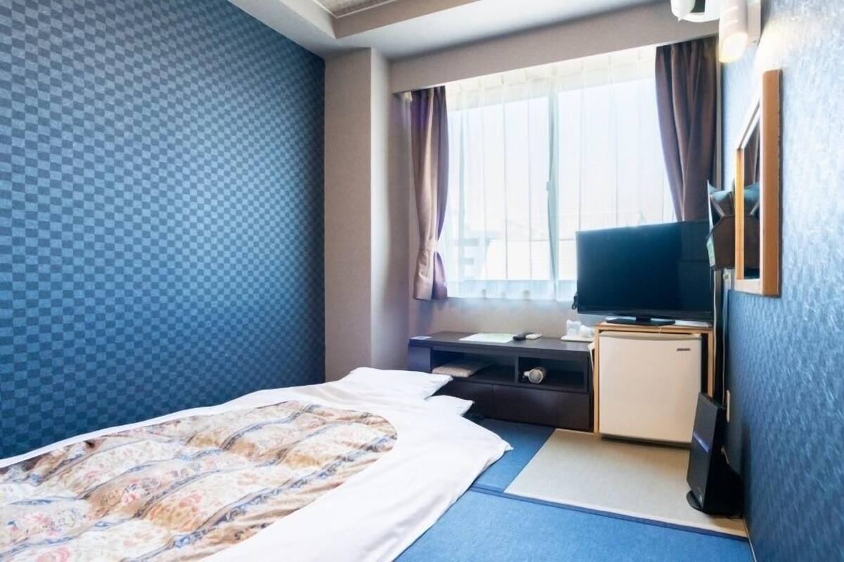 Quarto de hotel com cama, televisão, mini-frigorífico e janela com cortinas.