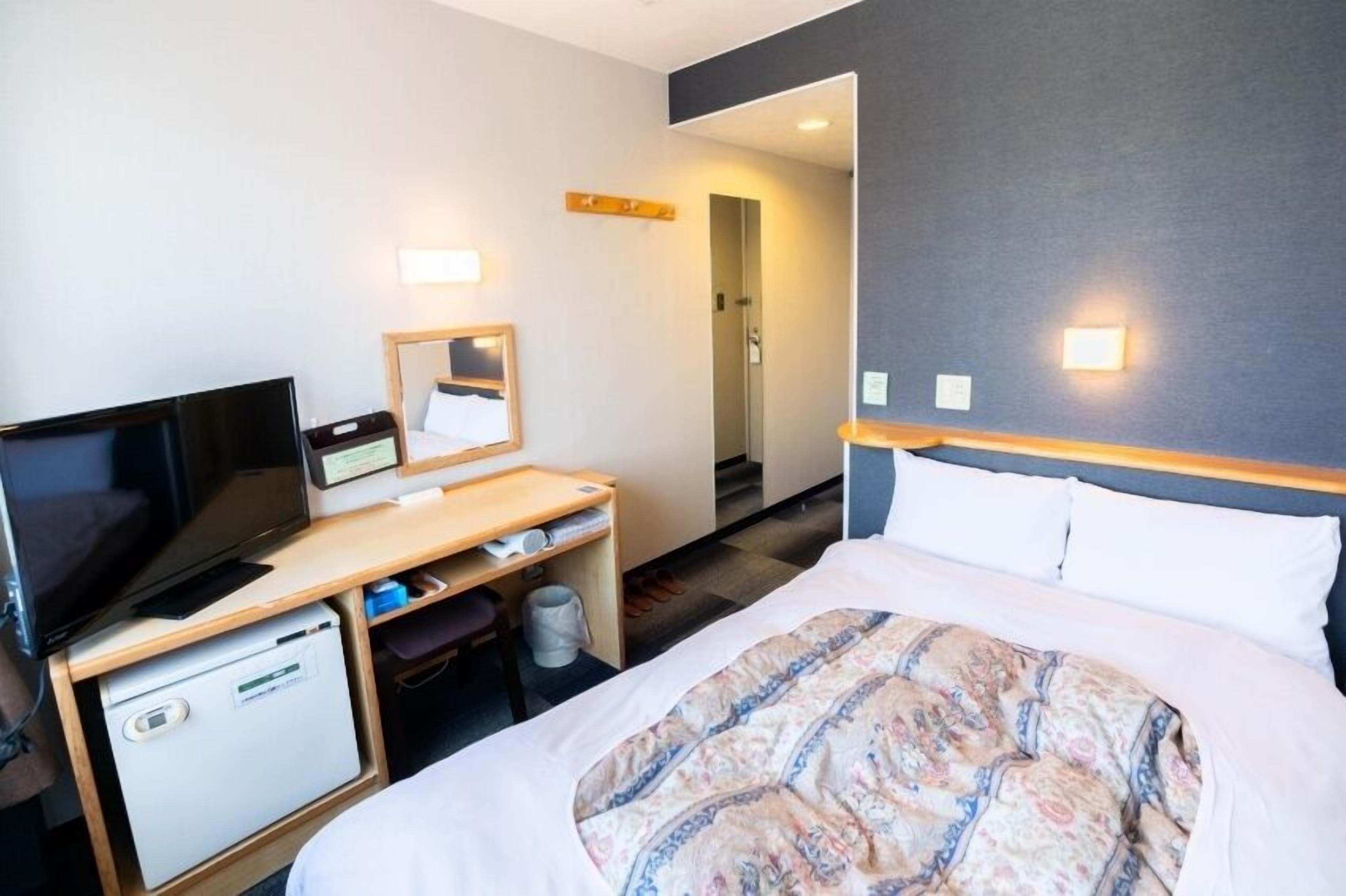 Quarto de hotel com uma cama, uma secretária com televisão, um espelho e um pequeno armário.