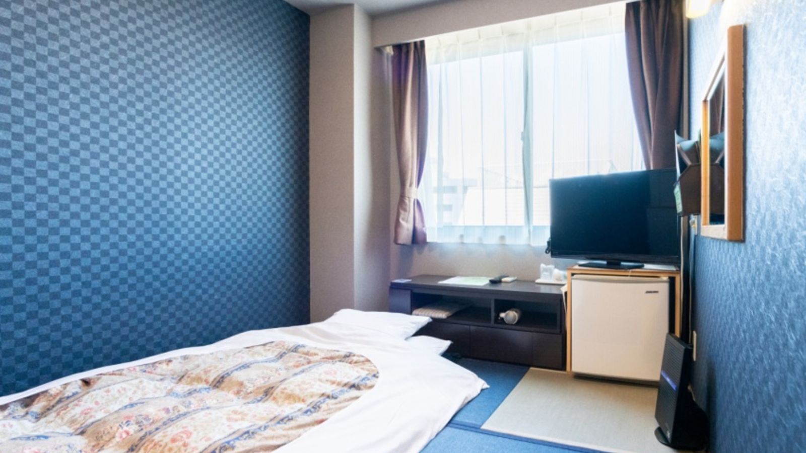 Quarto de hotel com uma cama, uma escrivaninha com televisão, um pequeno frigorífico e uma janela com cortinas.