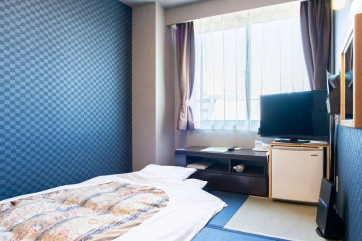 Quarto de hotel com uma cama, uma escrivaninha com televisão, um pequeno frigorífico e uma janela com cortinas.