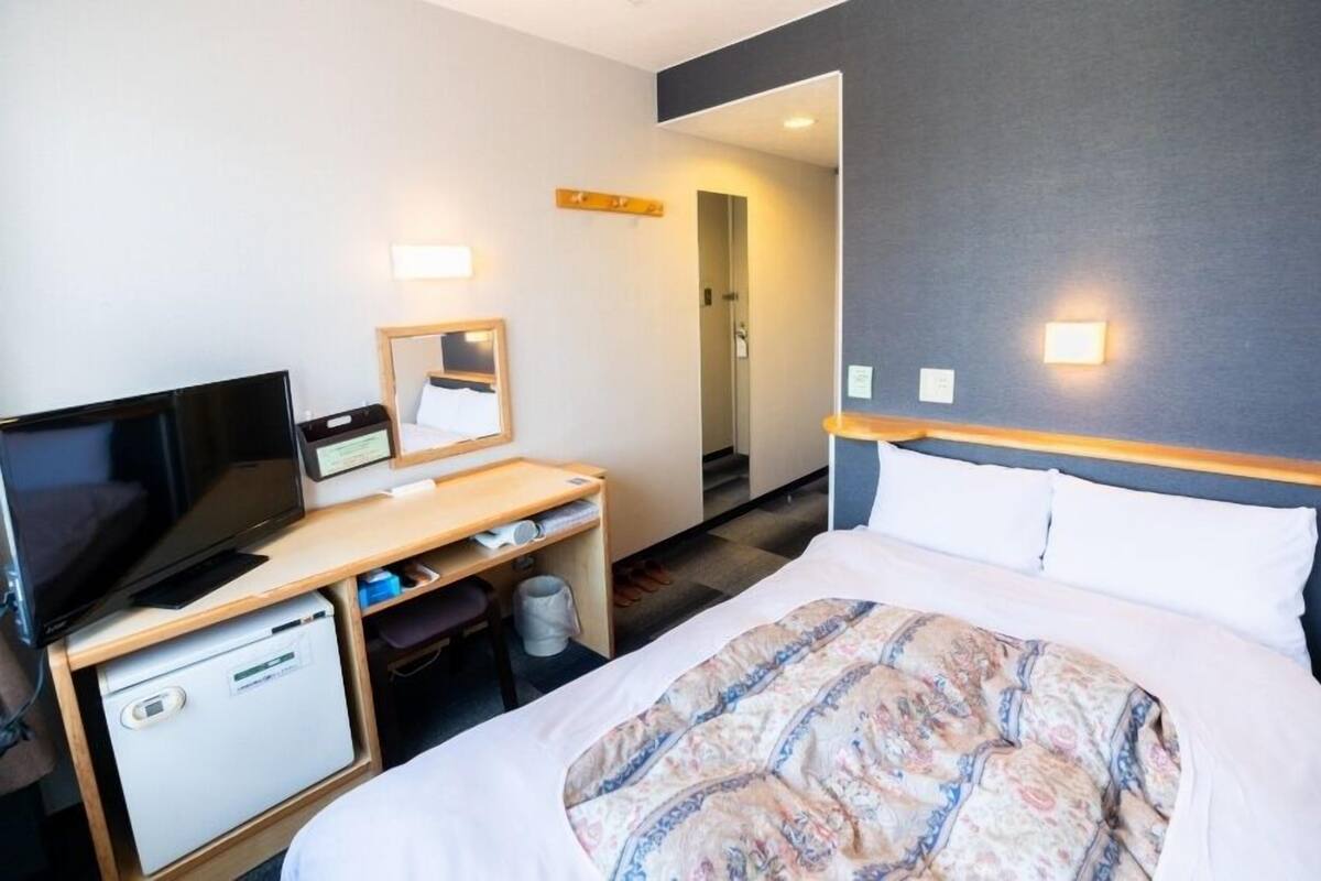Quarto de hotel com cama, escrivaninha, televisão, pequeno frigorífico e espelho.