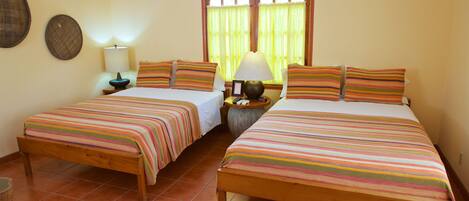 Quarto com duas camas de madeira, cada uma com roupa de cama listrada, uma mesa de cabeceira com abajur e uma janela com cortinas.