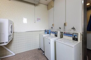 Uma área de lavanderia com duas máquinas de lavar e uma secadora, parede com azulejos e uma placa indicando a localização.