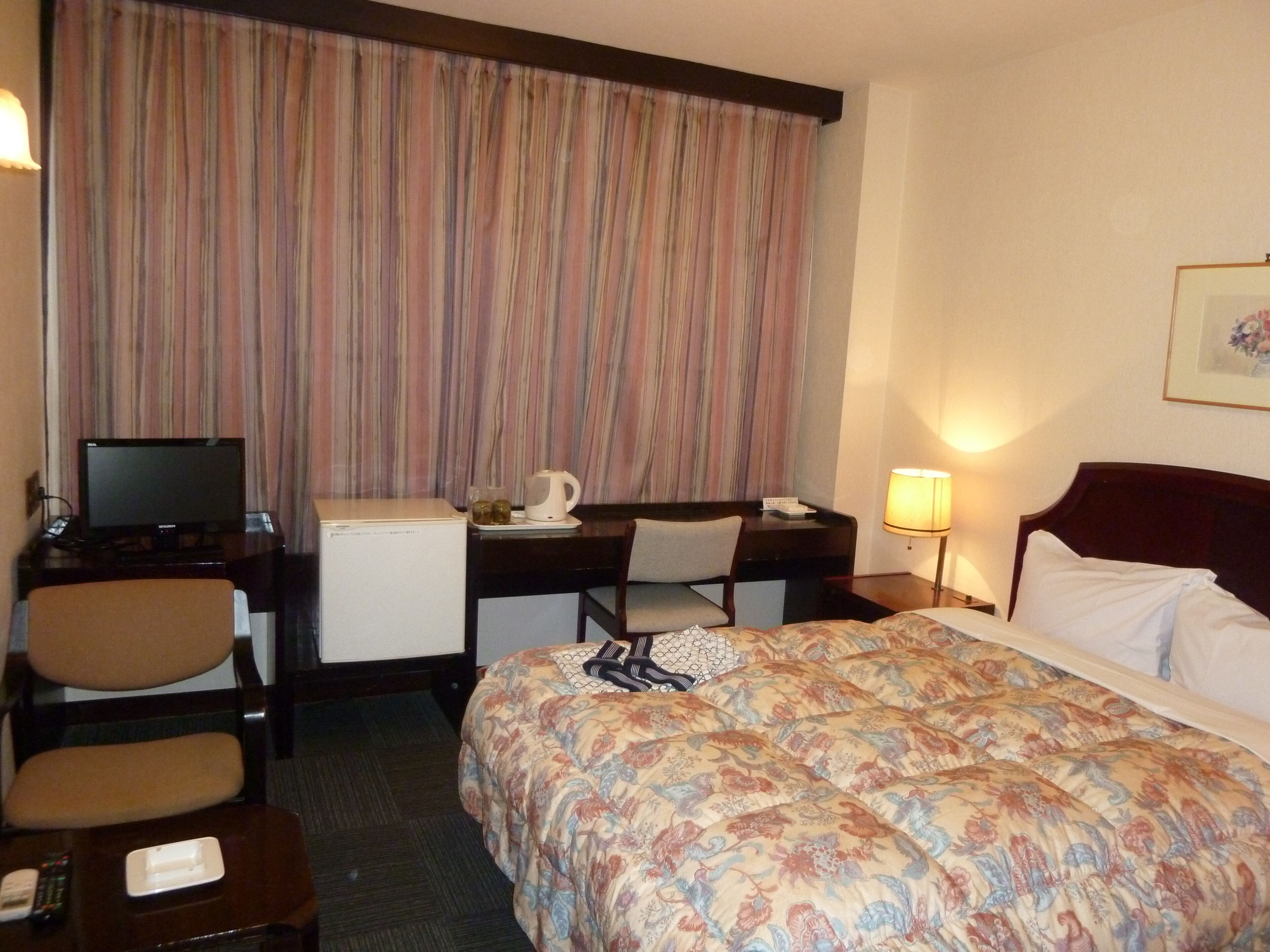 Quarto de hotel com cama, escrivaninha, cadeira, televisão e máquina de café.