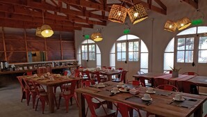 Uma ampla sala de jantar com mesas de madeira e cadeiras vermelhas, janelas grandes e luminárias pendentes.