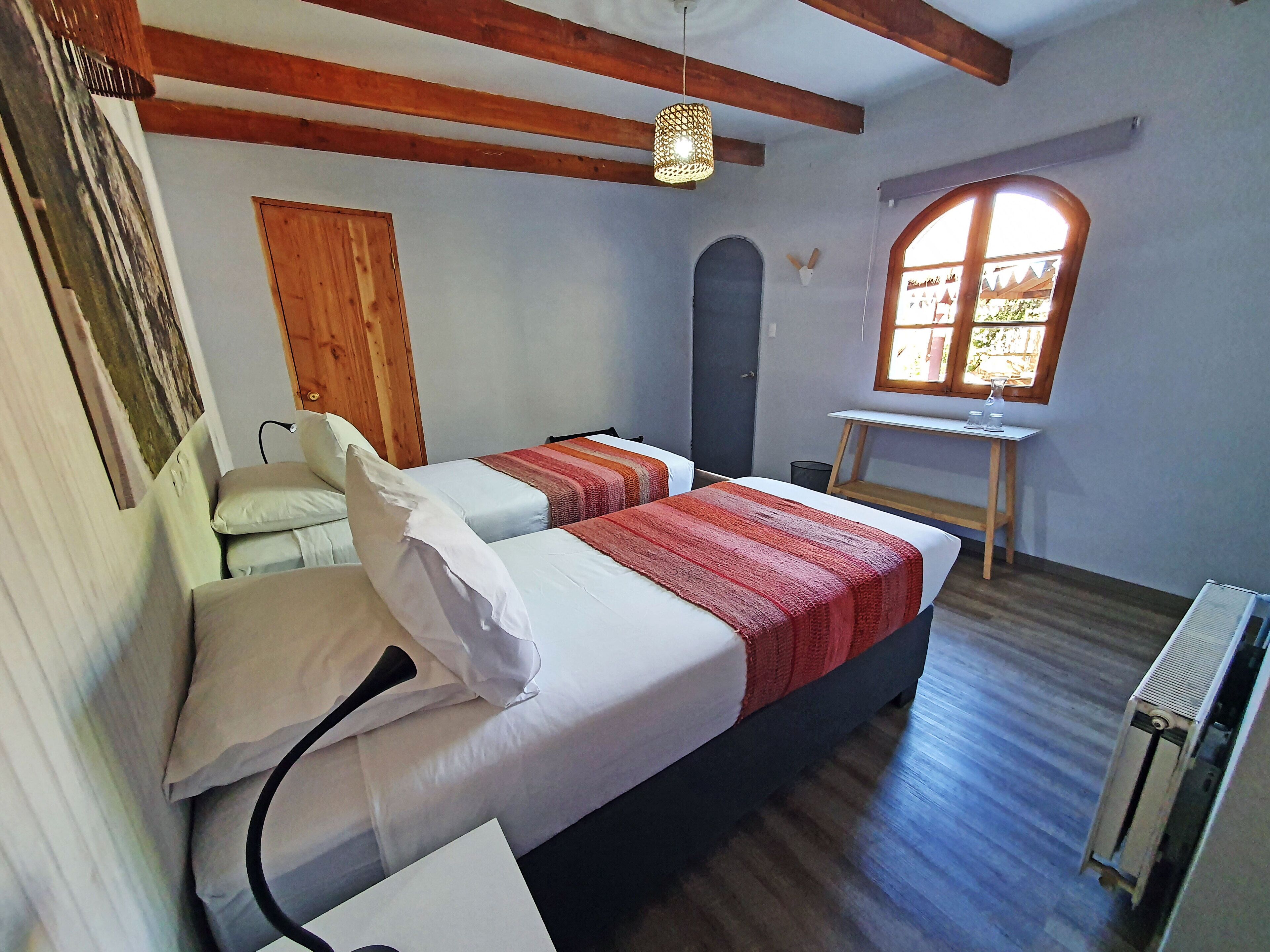 Quarto com duas camas, vigas de madeira, uma janela e uma pequena mesa.