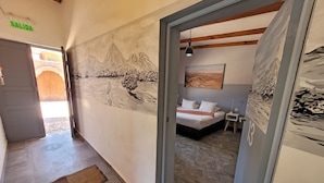 Corredor que dá acesso a um quarto com uma cama, uma mesinha de cabeceira e um quadro na parede.