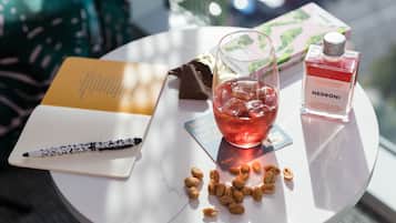 Uma mesa com um caderno, caneta, copo de bebida vermelha, frasco de perfume e alguns frutos secos espalhados.