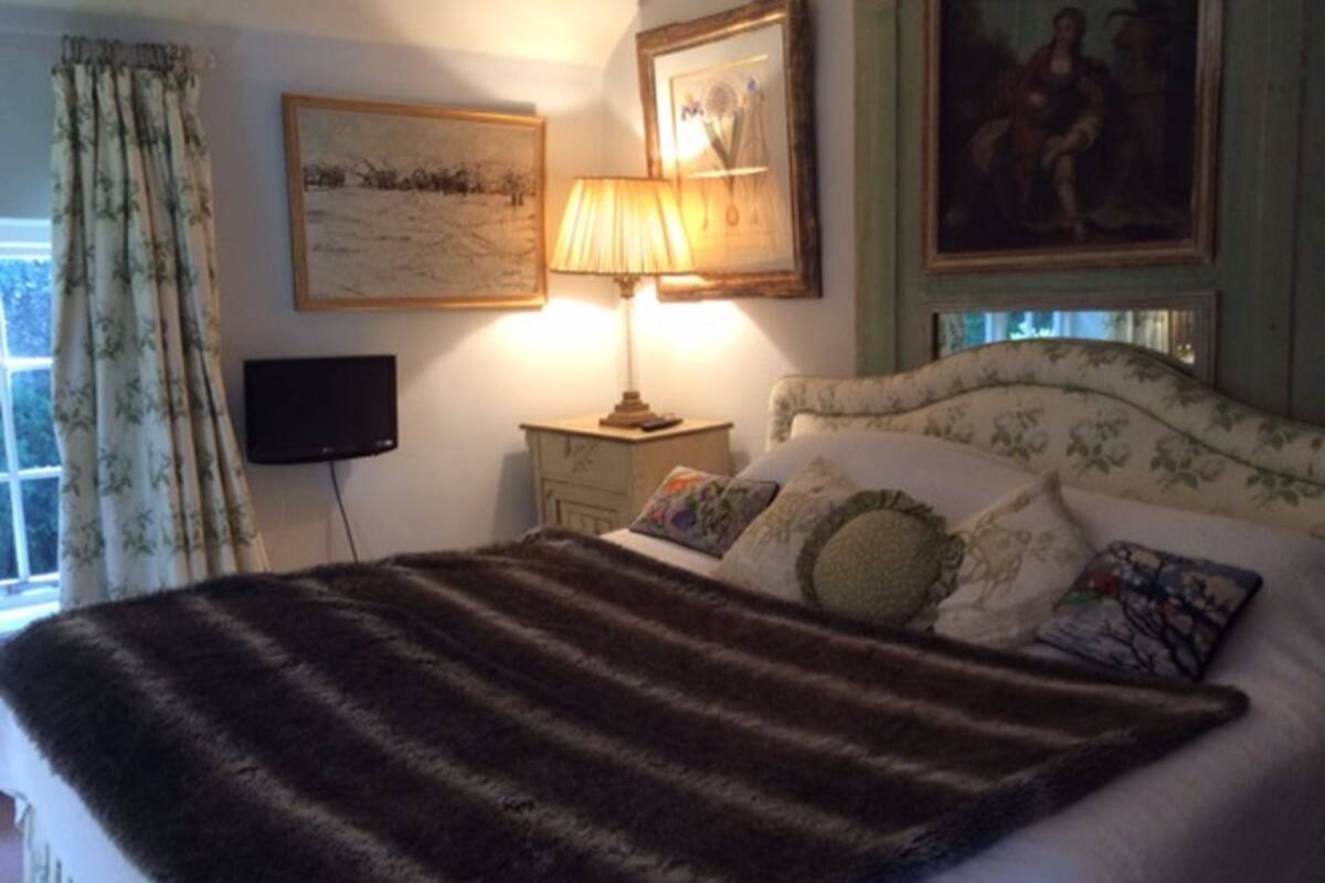 Quarto com cama, mesa de cabeceira, abajur, pintura e janela com cortinas.