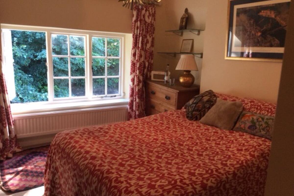 Quarto com uma janela grande, uma cama com roupa de cama estampada, cômoda e abajur.