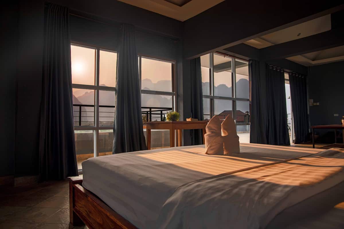 Quarto de hotel com uma cama grande, duas janelas com cortinas, uma mesinha com uma planta e vista para montanhas.