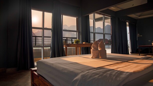 Quarto de hotel com uma cama grande, duas janelas com cortinas, uma mesinha com uma planta e vista para montanhas.
