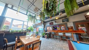 Restaurante com mesas e cadeiras de madeira, plantas penduradas e um espaço de bar.