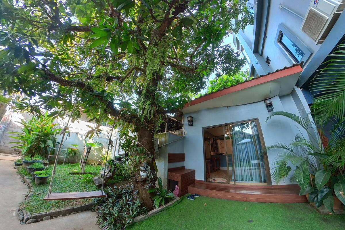 Um edifício residencial com um pequeno jardim, um deck de madeira e uma grande árvore que oferece sombra.