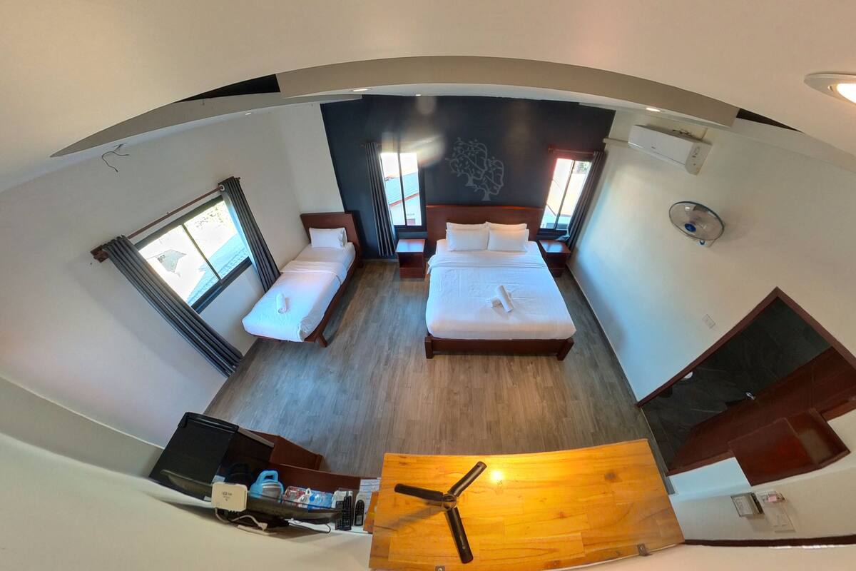 Vista panorâmica de um quarto de hotel com uma cama grande, duas camas menores, uma escrivaninha e uma cadeira.