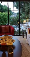 Quarto com uma grande janela, sofá vermelho, mesa com jogos americanos trançados e vista para uma piscina e vegetação.