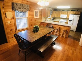 Interior em madeira com mesa de jantar, cadeiras, um banco e uma cozinha com armários e eletrodomésticos.
