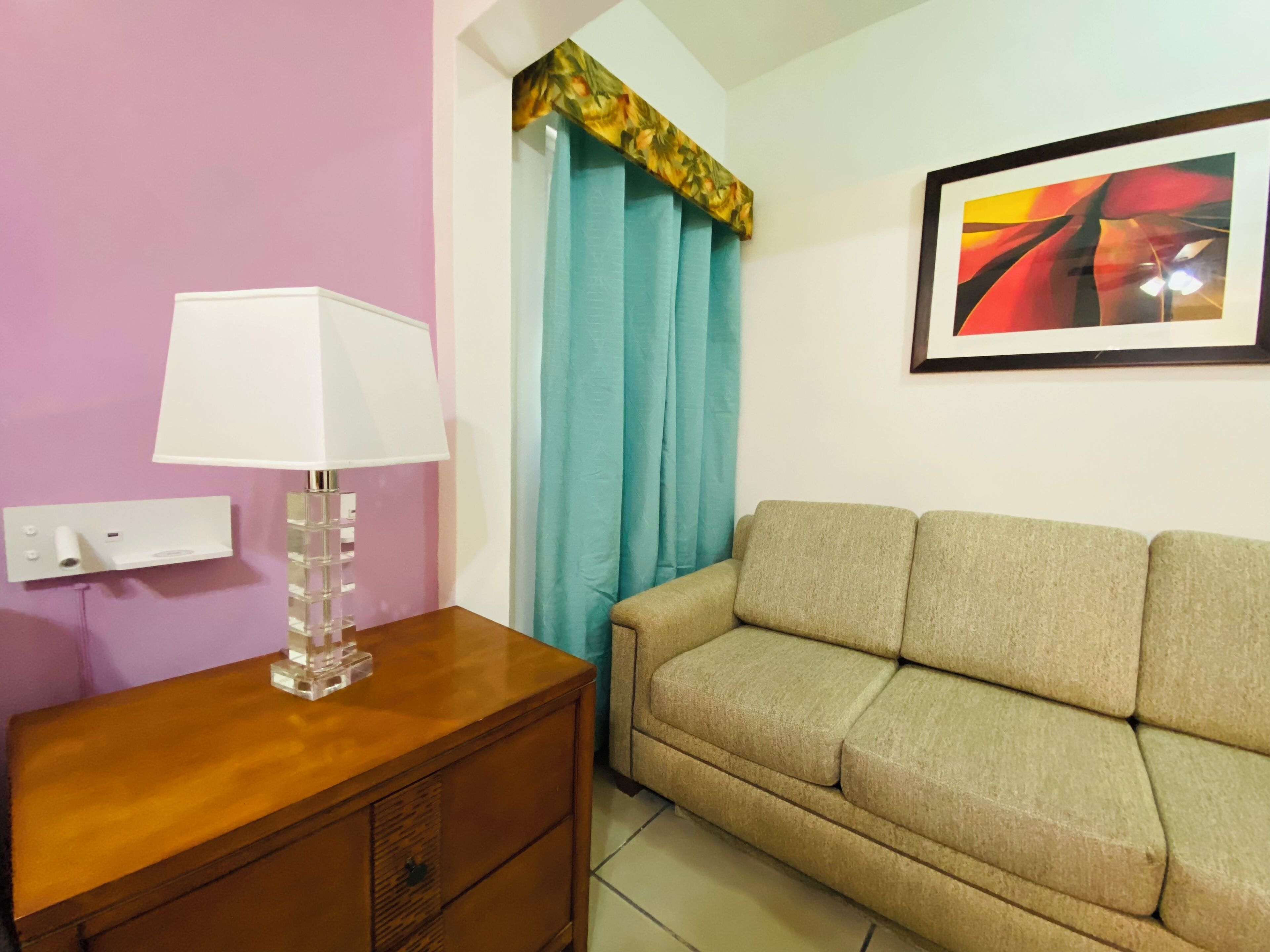 Quarto com mesa de cabeceira de madeira, luminária, sofá bege, tomada na parede e pintura abstrata emoldurada.