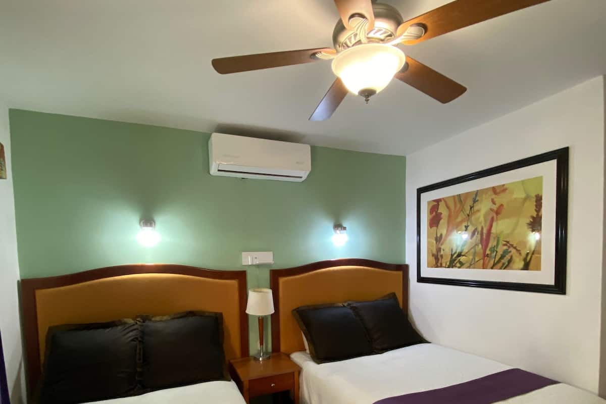 Quarto de hotel com duas camas, ventoinha no teto, ar condicionado e uma moldura na parede.