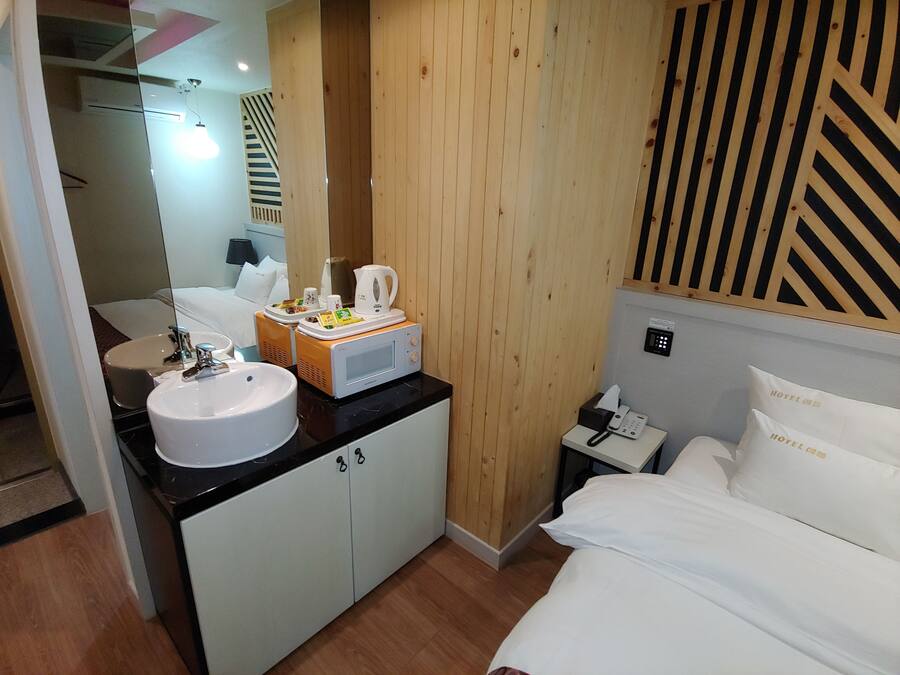 Quarto de hotel com parede de madeira, uma cama, pia branca, micro-ondas e mesa de cabeceira com telefone e lenços.
