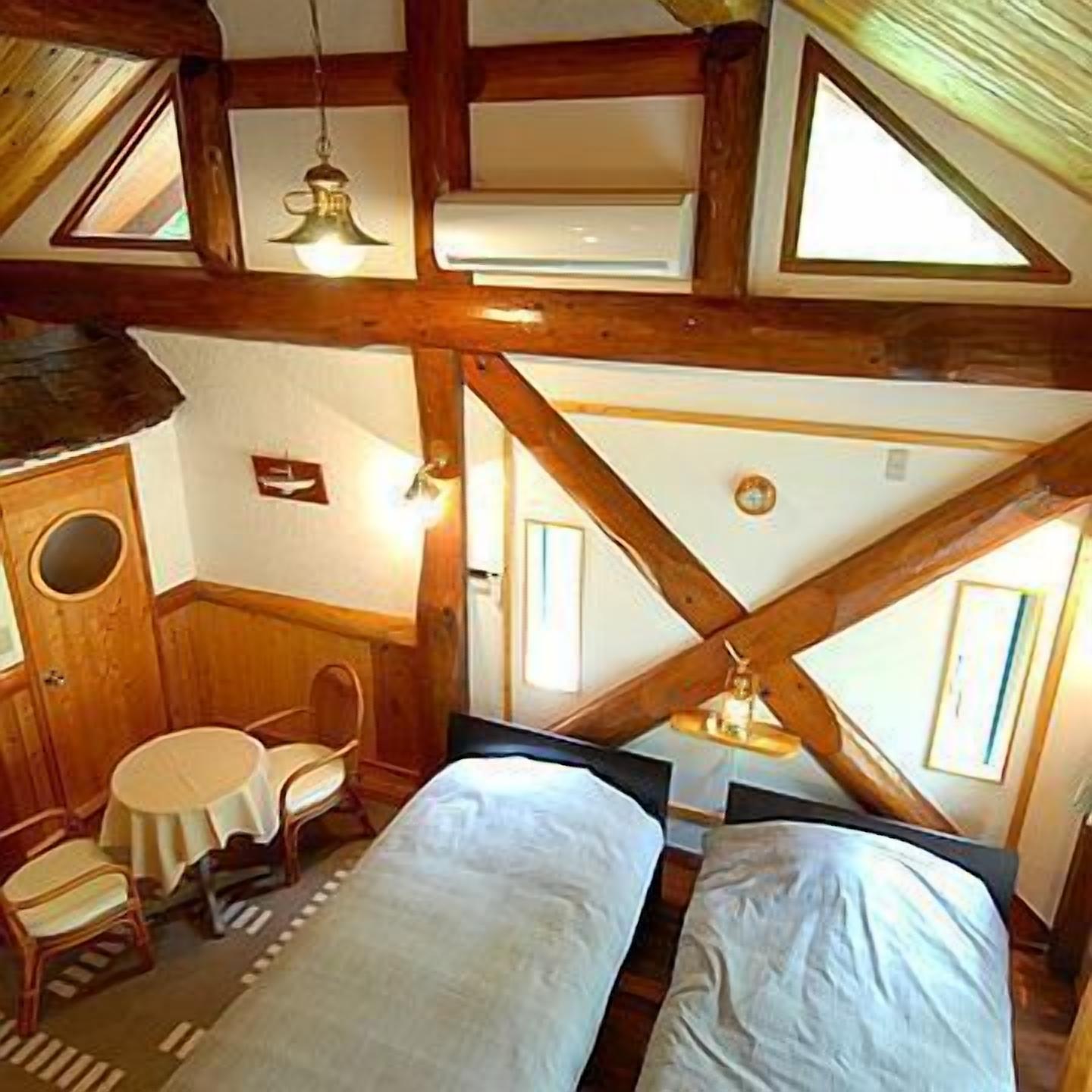 Quarto com teto inclinado, vigas de madeira, duas camas simples, uma mesa redonda pequena, uma cadeira e uma janela.