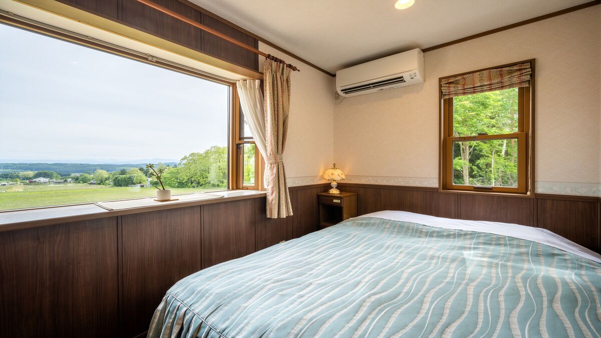 Quarto com uma grande janela, cama com colcha estampada, mesa de cabeceira com abajur e ar condicionado na parede.