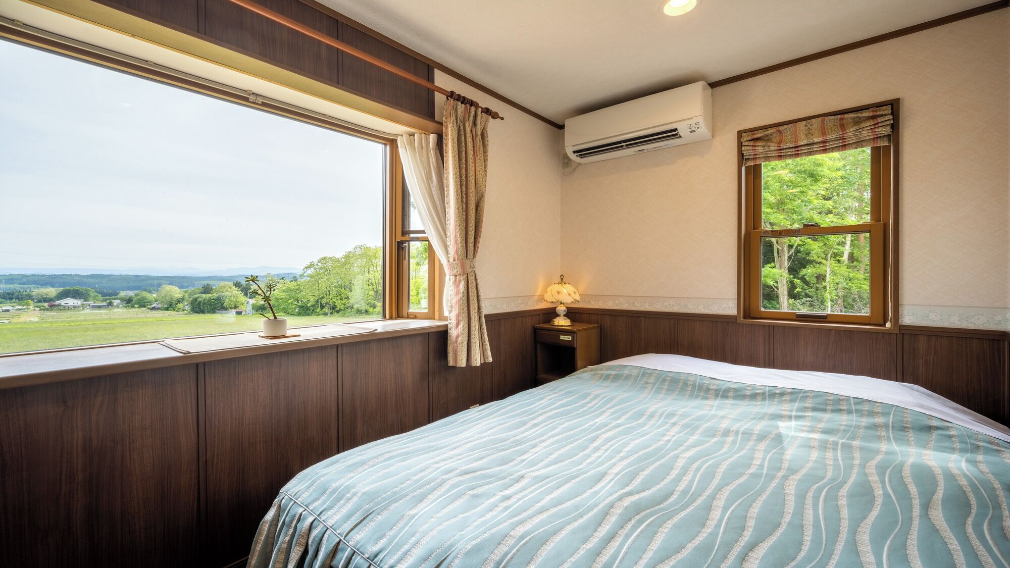 Quarto com cama, mesa de cabeceira, ar condicionado, janela com cortinas e vista para o verde.
