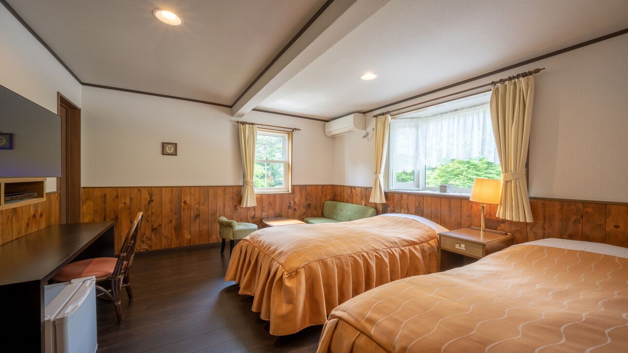 Quarto de hotel com duas camas, uma mesa, uma cadeira, janela com cortinas e um aparelho de ar condicionado.