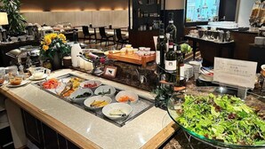 Disposição de buffet com diversos alimentos, seleção de vinhos e uma saladeira.