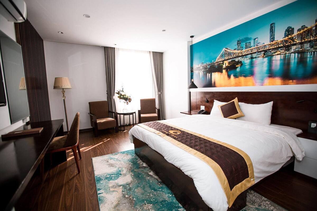 Quarto de hotel com uma cama grande, uma mesa, uma cadeira, um abajur e um quadro com vista para a cidade.