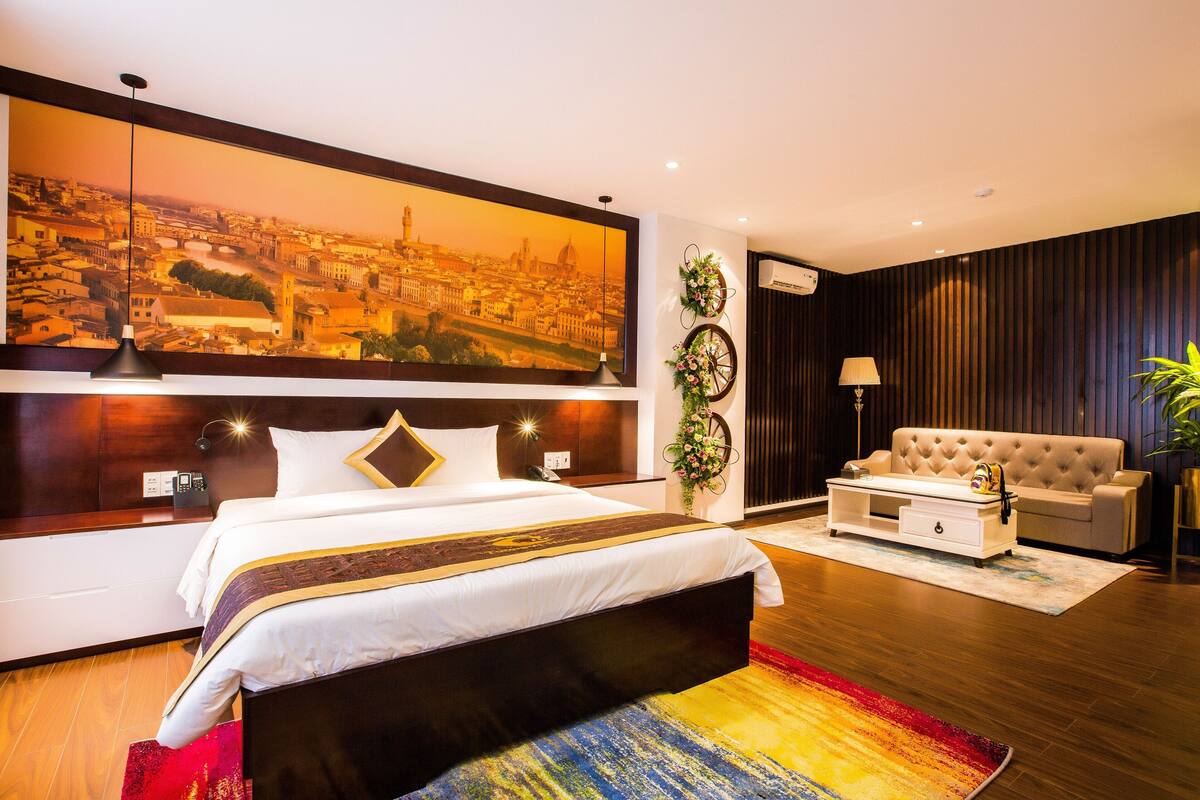 Quarto de hotel moderno com uma cama grande, um sofá, uma mesa de centro e uma pintura de paisagem urbana na parede.