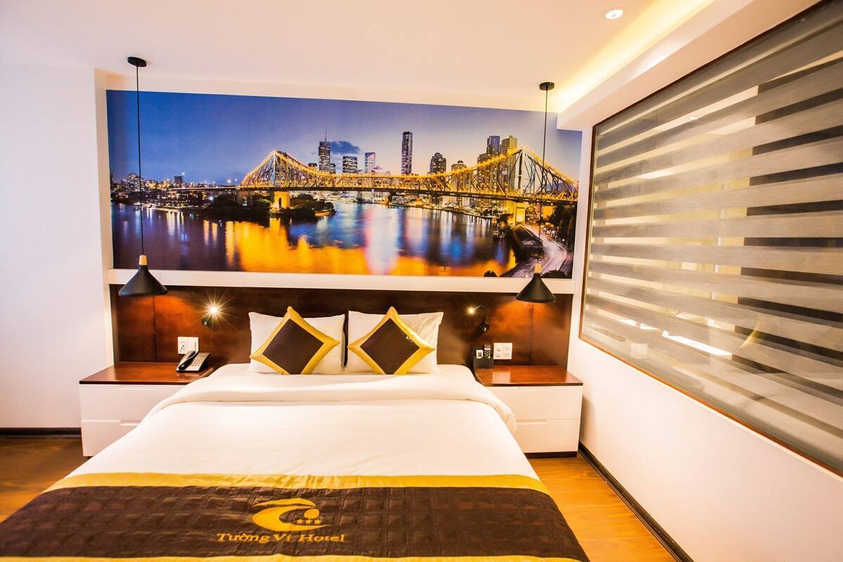 Quarto de hotel com cama, mesas de cabeceira, um mural da paisagem urbana e uma janela com persianas.