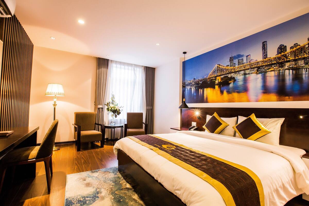 Quarto de hotel com uma cama grande, uma escrivaninha, uma cadeira, um abajur e uma pintura de paisagem urbana na parede.