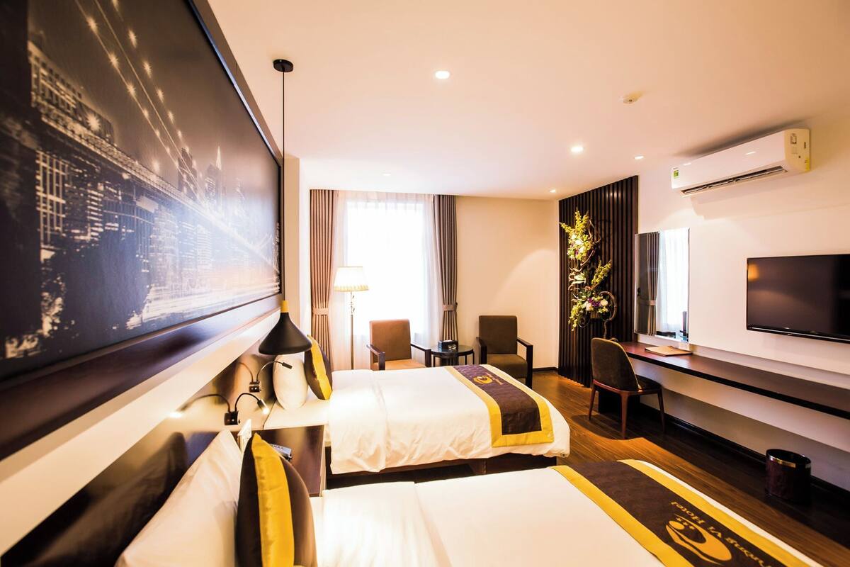 Quarto de hotel com duas camas, uma escrivaninha, televisão e uma janela grande com cortinas.