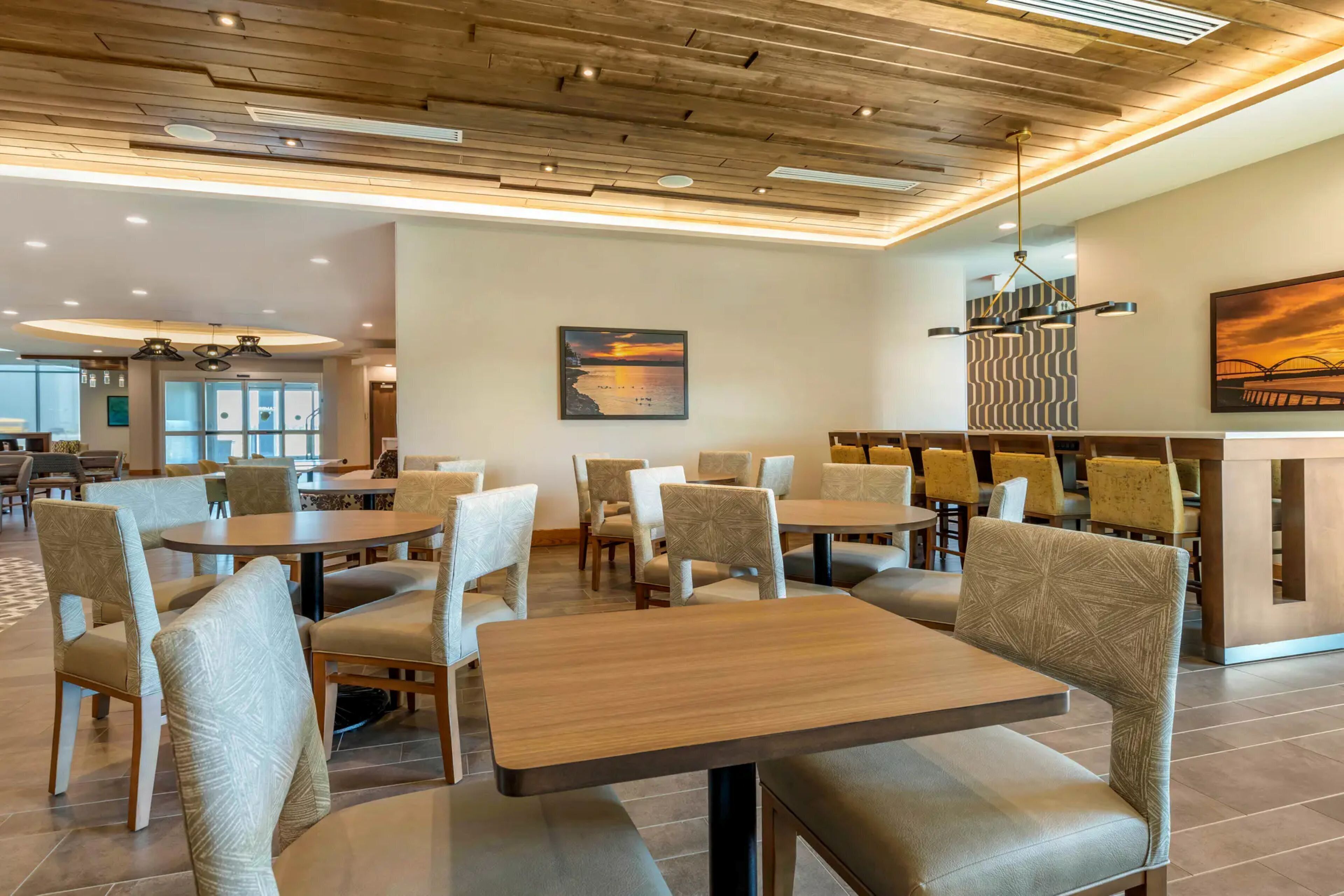 Um restaurante moderno com mesas redondas e cadeiras estofadas, teto de madeira e obras de arte nas paredes.