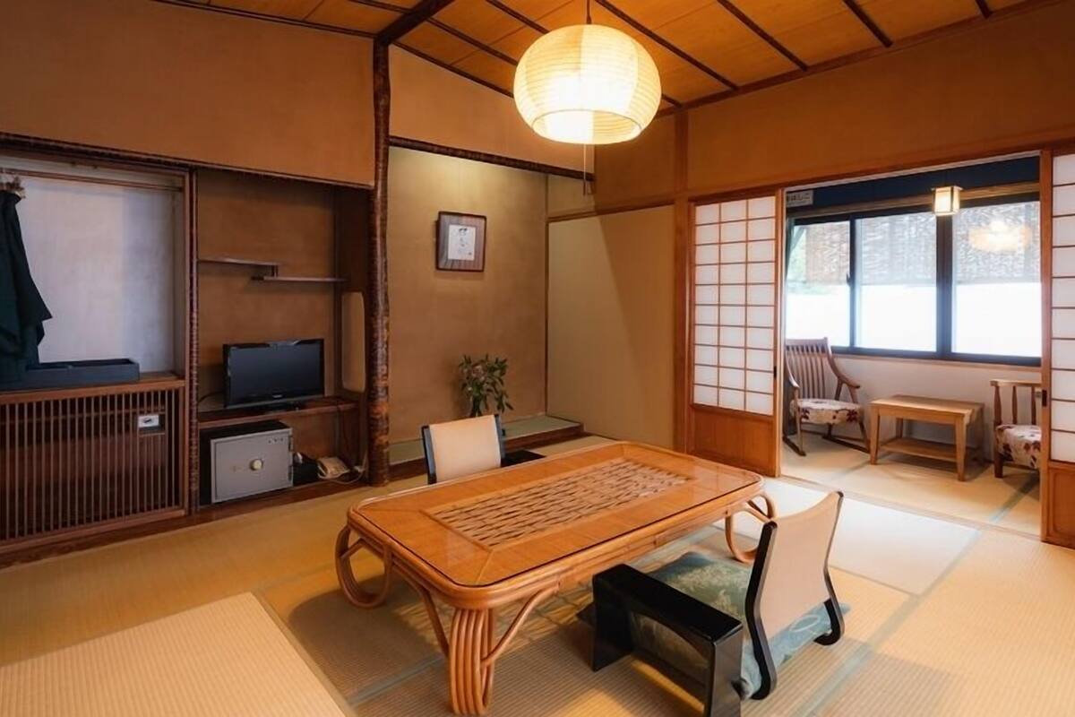 Um quarto japonês tradicional com piso de tatame, uma mesa baixa, cadeiras, uma televisão e portas deslizantes.